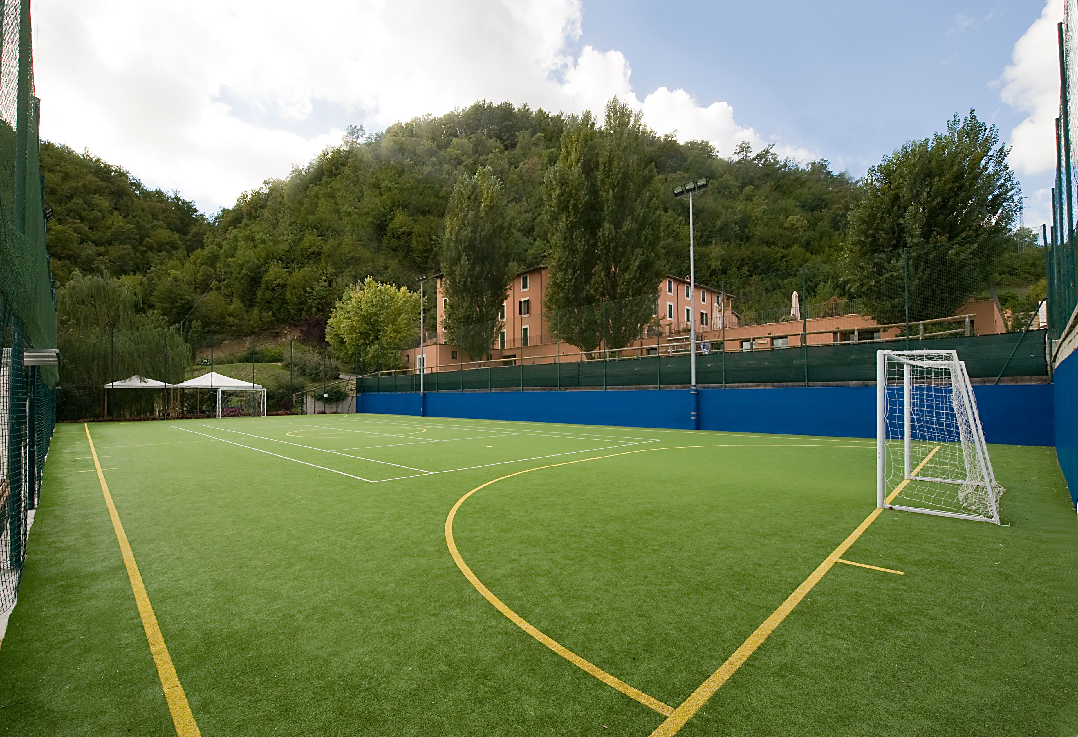 Um campo de futebol coberto com um gol, cercado por uma grade verde, e um edifício ao fundo.
