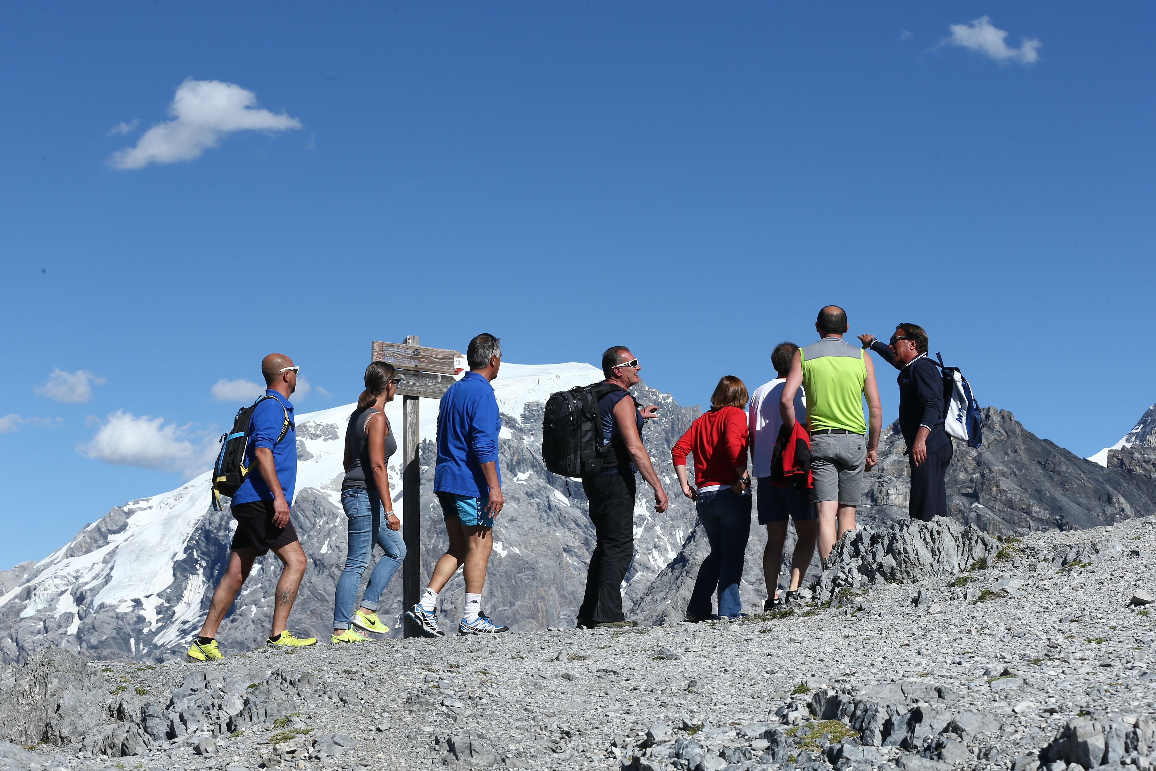 Um grupo de caminhantes em uma trilha rochosa na montanha, com picos nevados ao fundo.