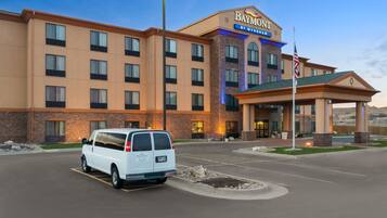 Hotel Baymont by Wyndham com uma carrinha branca estacionada em frente, uma entrada bem iluminada e uma bandeira hasteada.