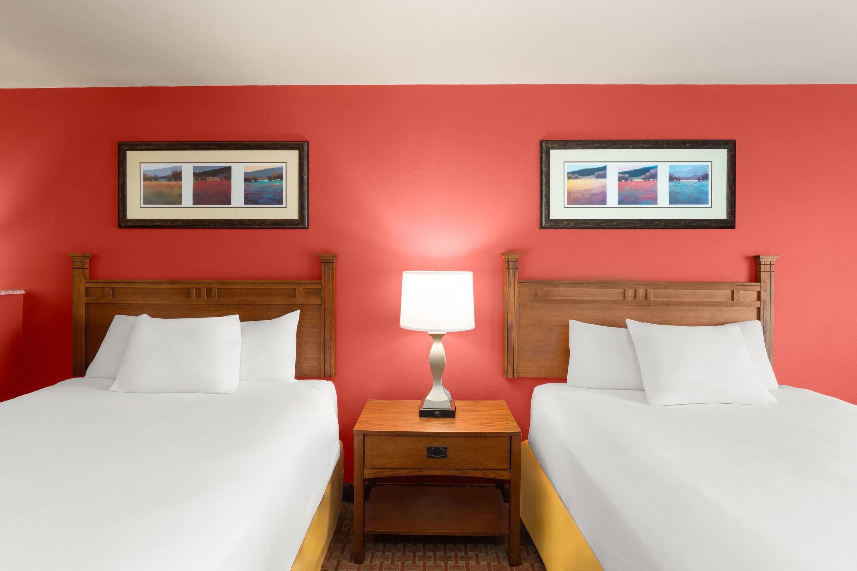 Quarto de hotel com duas camas, uma mesa de cabeceira com abajur e duas fotografias emolduradas na parede.