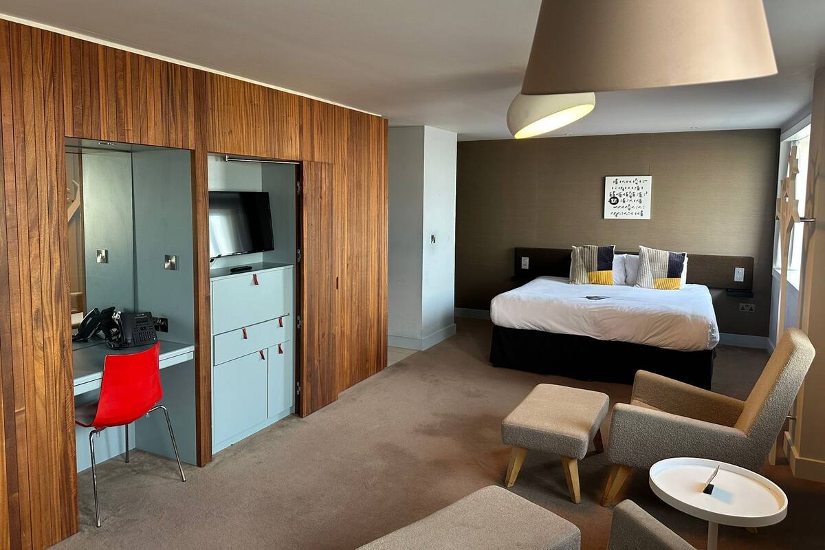 Quarto de hotel com cama, escrivaninha com cadeira, pequena mesa e guarda-roupa.