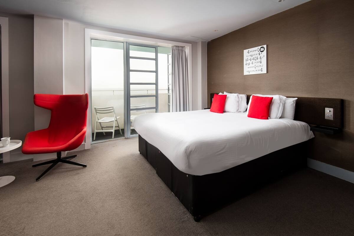 Quarto de hotel com uma cama grande, uma cadeira vermelha, uma mesinha e uma porta de vidro deslizante para a varanda.