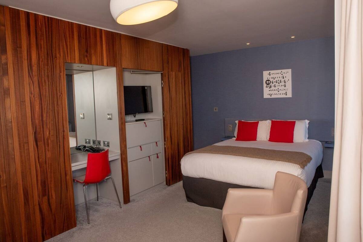 Quarto de hotel com cama, escrivaninha, cadeira, televisão e espelho.