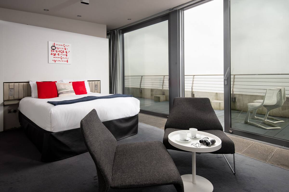 Quarto de hotel moderno com uma cama grande, duas poltronas, uma mesa pequena com bandeja e uma varanda com cadeira.