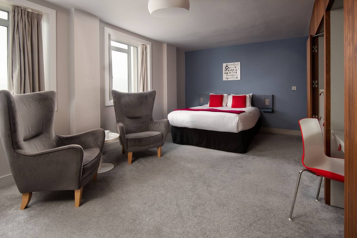 Quarto de hotel com uma cama, duas poltronas, uma mesinha e uma cadeira vermelha.
