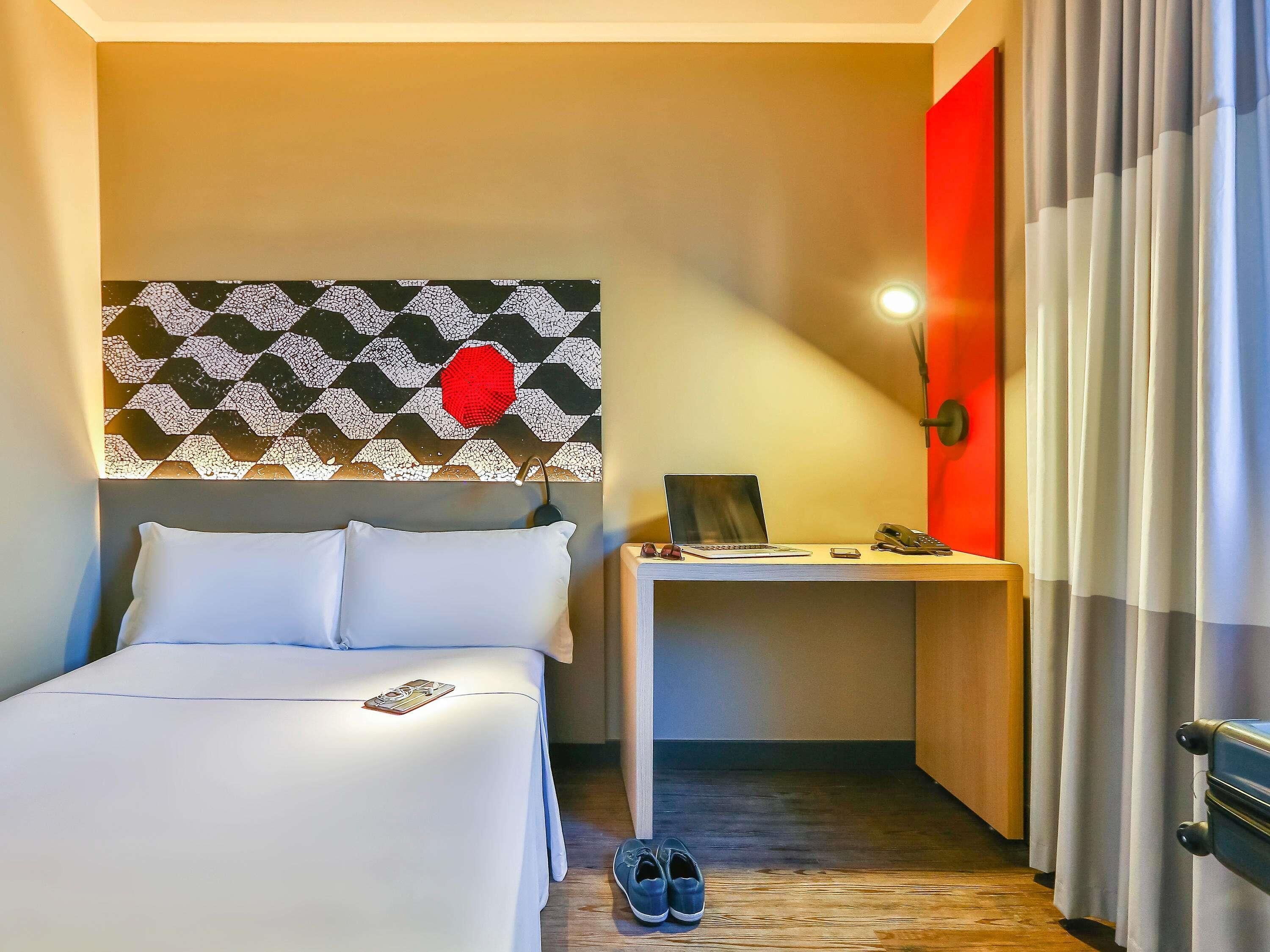 Quarto de hotel com cama, uma secretária com um computador portátil, telefone e um par de sapatos no chão.