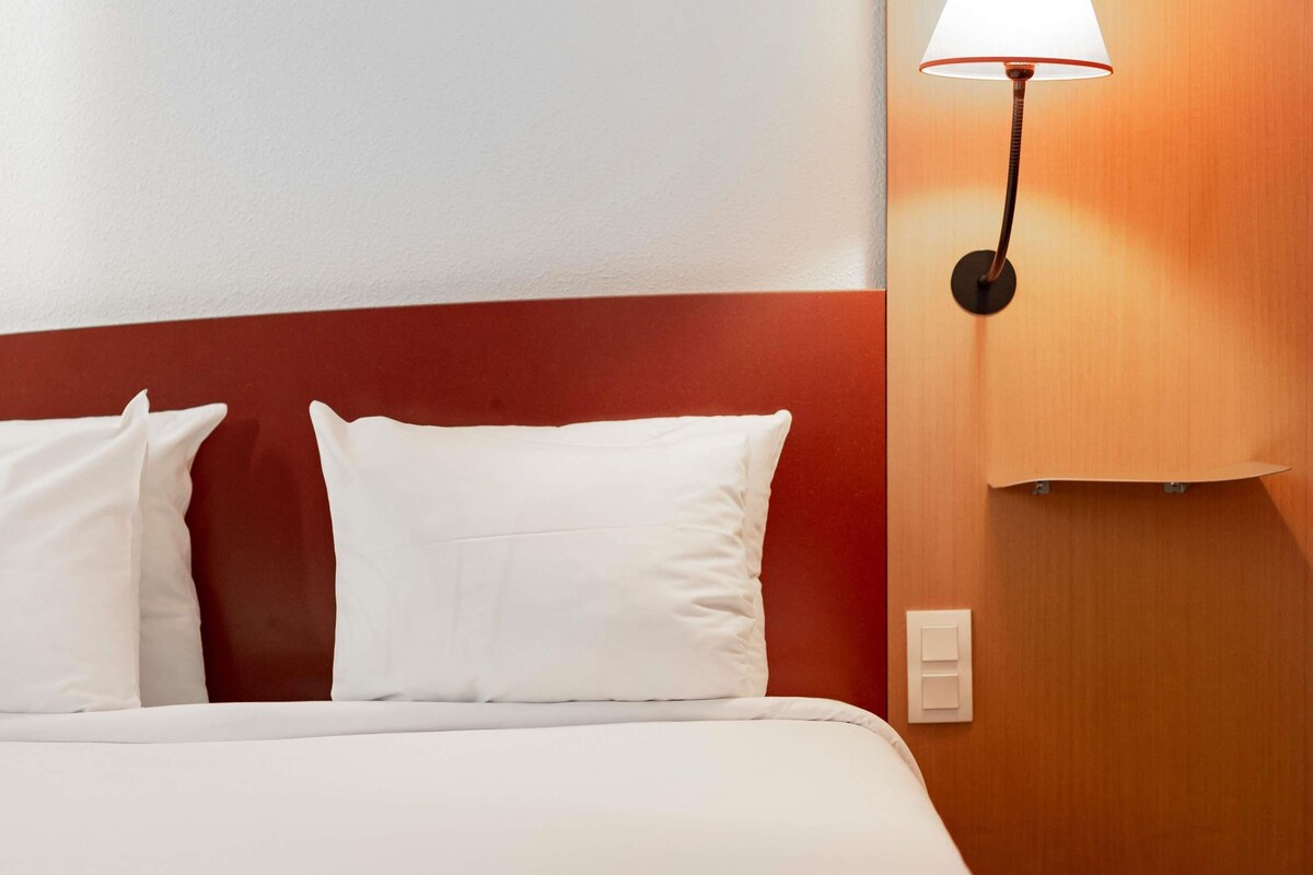 Quarto de hotel com cama, almofadas brancas, cabeceira de madeira e luminária na parede.