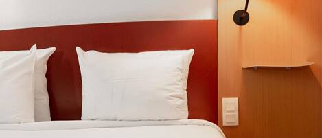 Quarto de hotel com cama, almofadas brancas, cabeceira de madeira e luminária na parede.