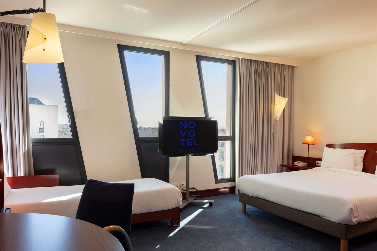 Quarto de hotel com duas camas, uma escrivaninha, televisão e janelas amplas.
