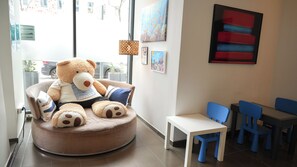 Quarto com um urso de pelúcia grande em uma cadeira redonda, uma mesinha, cadeiras azuis e obras de arte nas paredes.