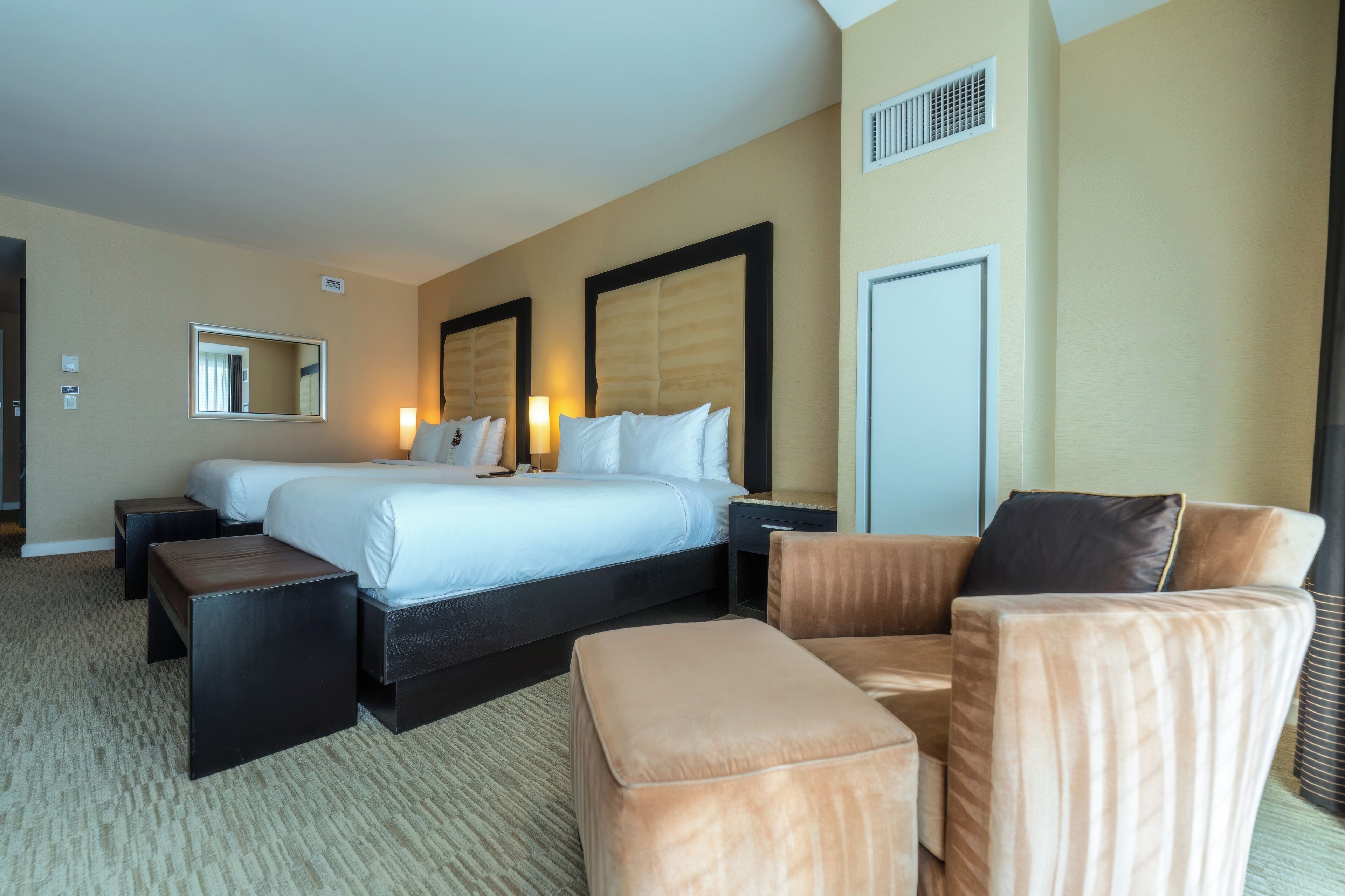 Quarto de hotel com uma cama grande, duas mesas de cabeceira, uma cadeira e um pequeno banquinho.