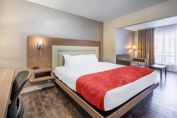 Quarto de hotel com cama, escrivaninha, cadeira, mesa de cabeceira, luminária e janela com cortinas.