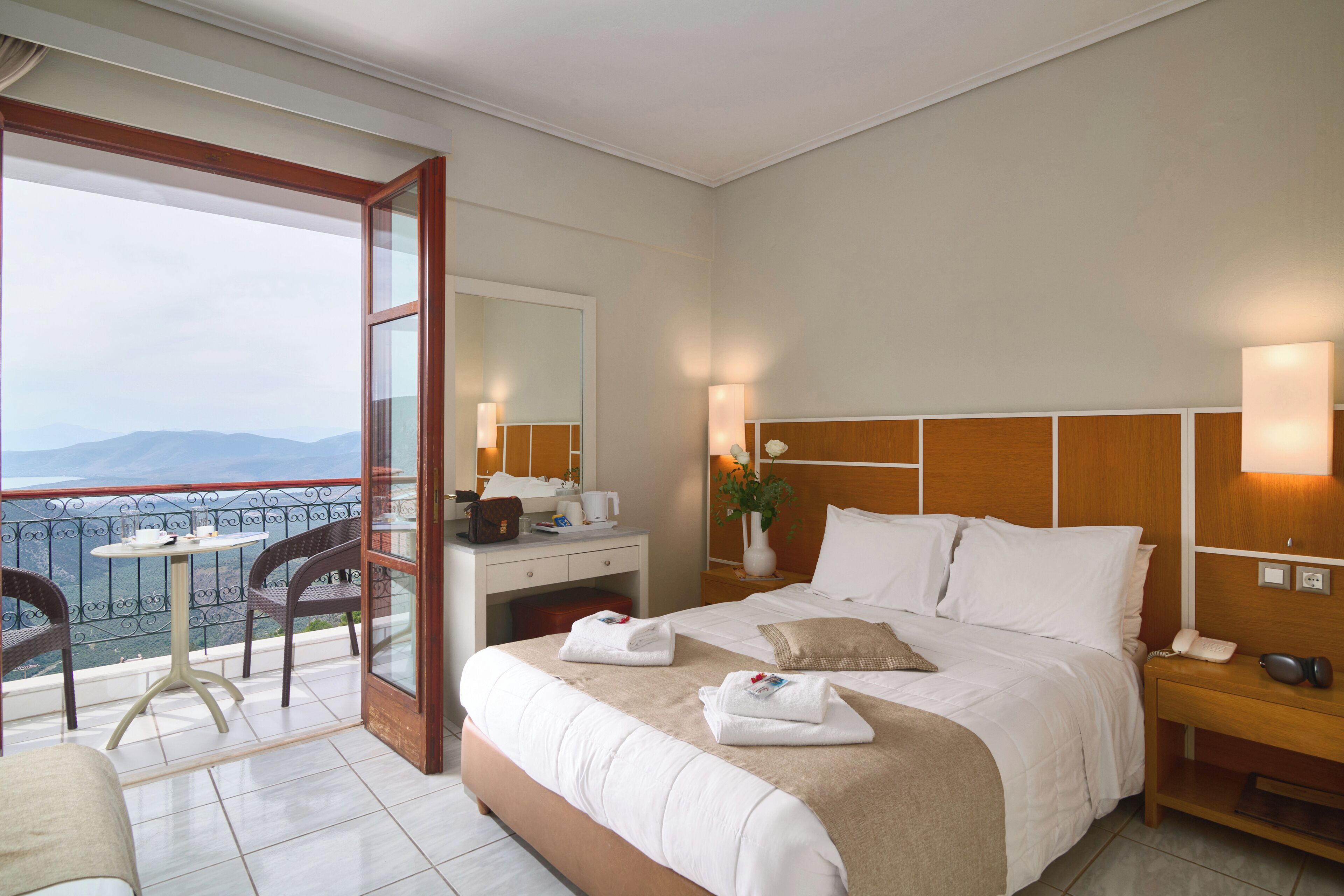 Quarto de hotel com cama, mesa de cabeceira, e varanda com mesa e cadeiras.