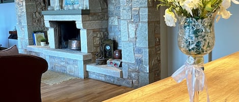 Uma lareira de pedra com um relógio e objetos decorativos, uma mesa de centro de madeira e um vaso com flores.
