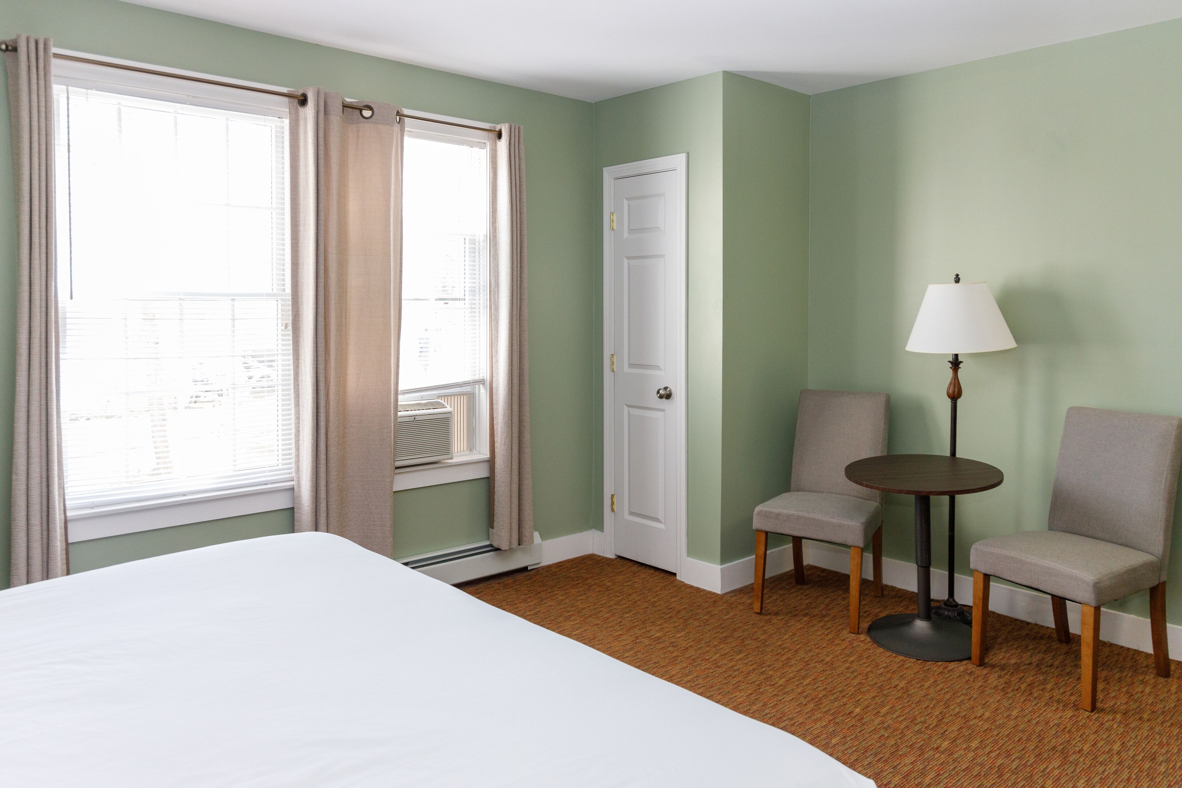 Quarto de hotel com cama, duas cadeiras, uma mesa, um abajur e uma porta.