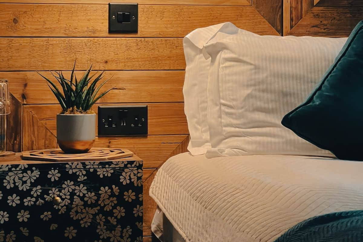 Um quarto aconchegante com cabeceira de madeira, mesa de cabeceira com uma planta, abajur e cama com roupa de cama branca e almofada verde-água.