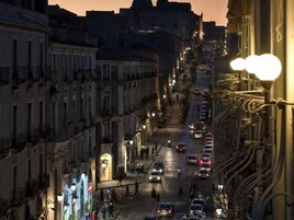 Vista da rua ao anoitecer, com edifícios em ambos os lados, carros na estrada e postes de luz.