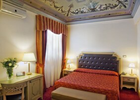Quarto de hotel com cama, duas mesas de cabeceira com abajures, escrivaninha com luminária, cadeira e decoração no teto.