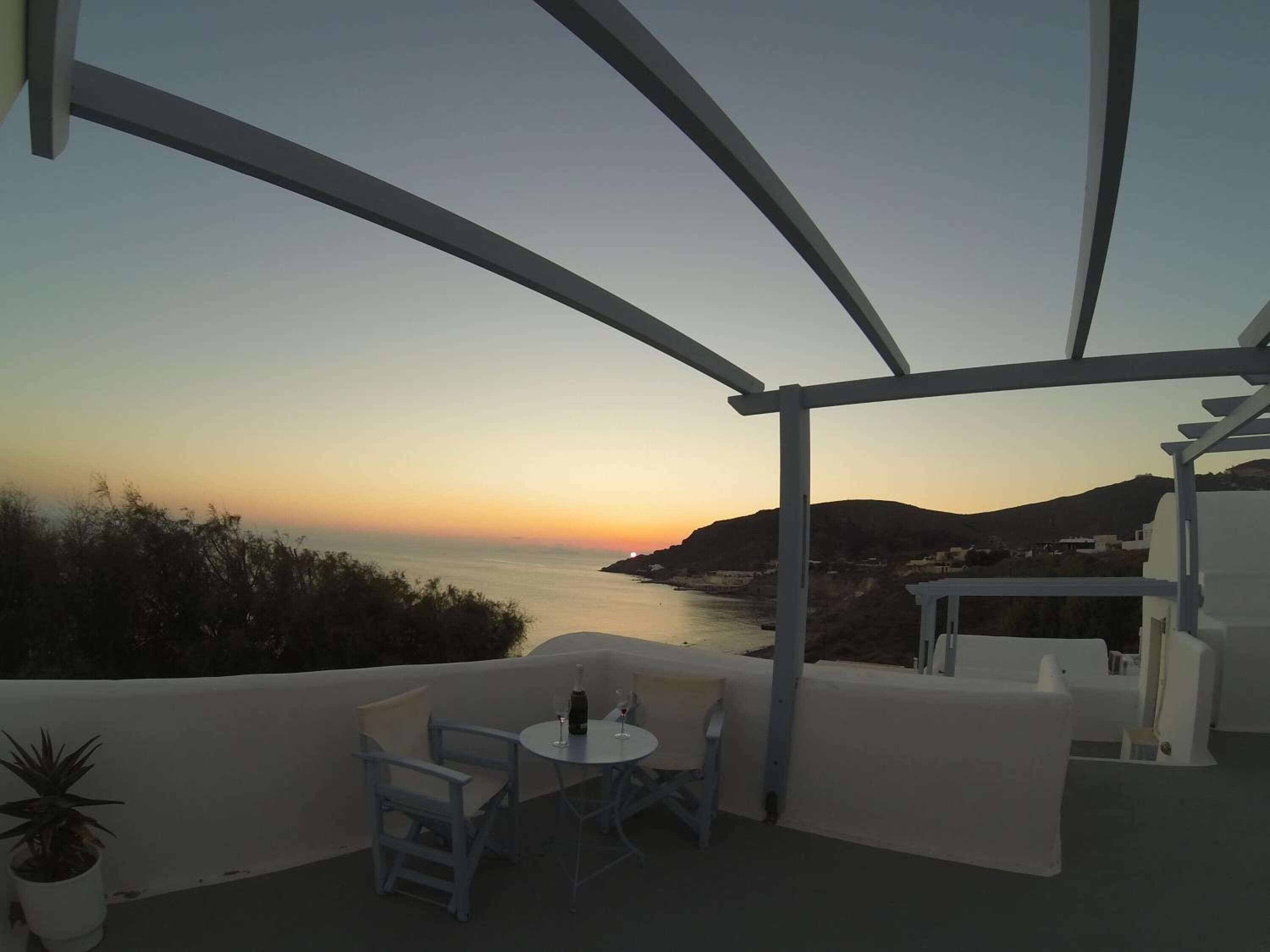 Varanda com mesa e cadeiras, vista para a costa com um pôr do sol.