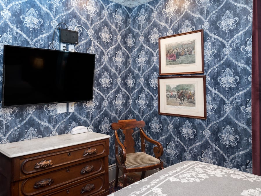 Quarto com uma grande TV de tela plana na parede, uma cadeira de madeira com assento acolchoado, uma cômoda de madeira com gavetas e duas imagens emolduradas penduradas na parede.