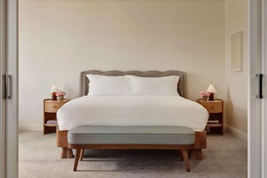 Cama bem arrumada com cabeceira de madeira, duas mesas de cabeceira com luminárias e um banco ao pé da cama.