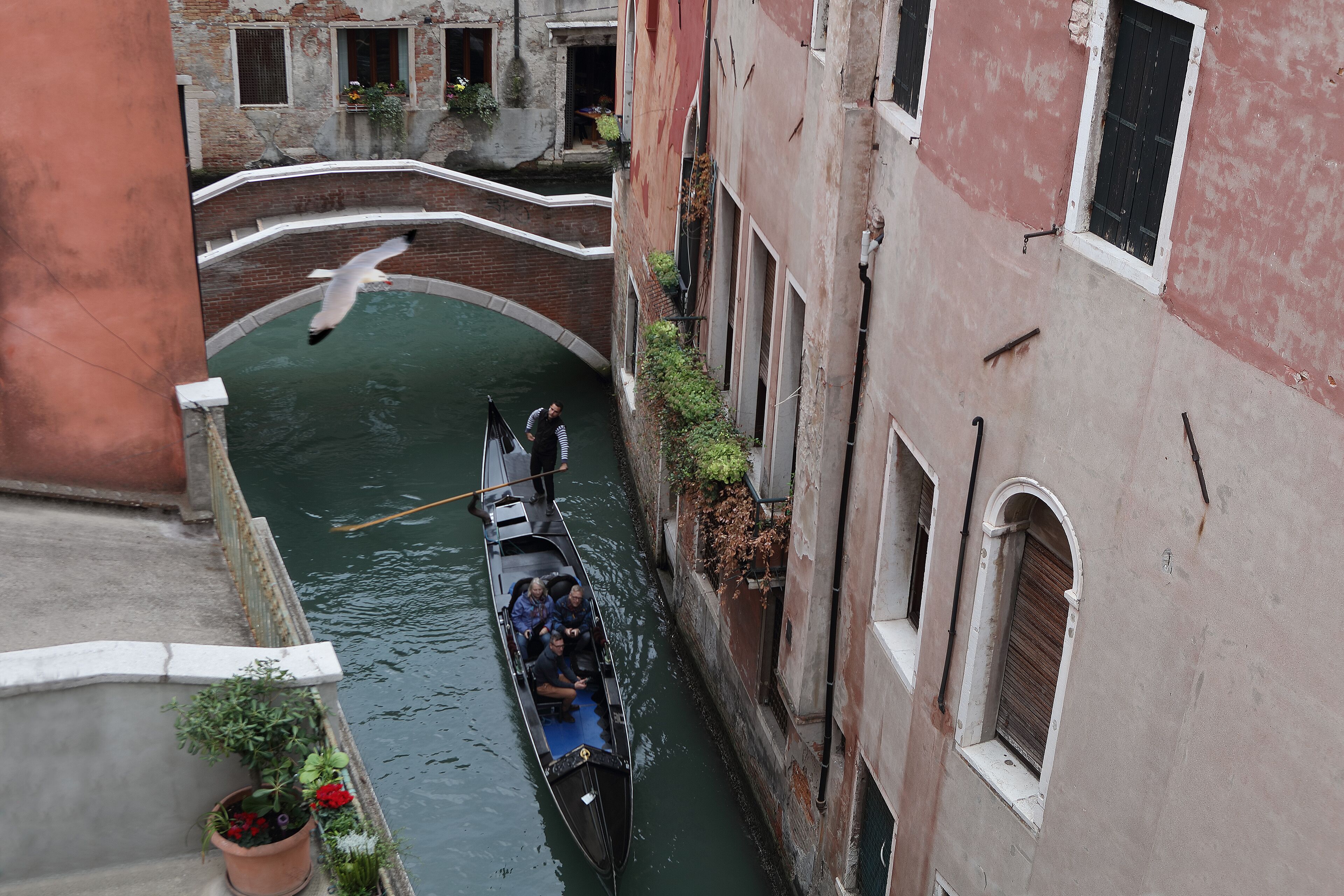 Um canal estreito com uma gôndola transportando passageiros, ladeado por edifícios históricos com janelas em arco e varandas.