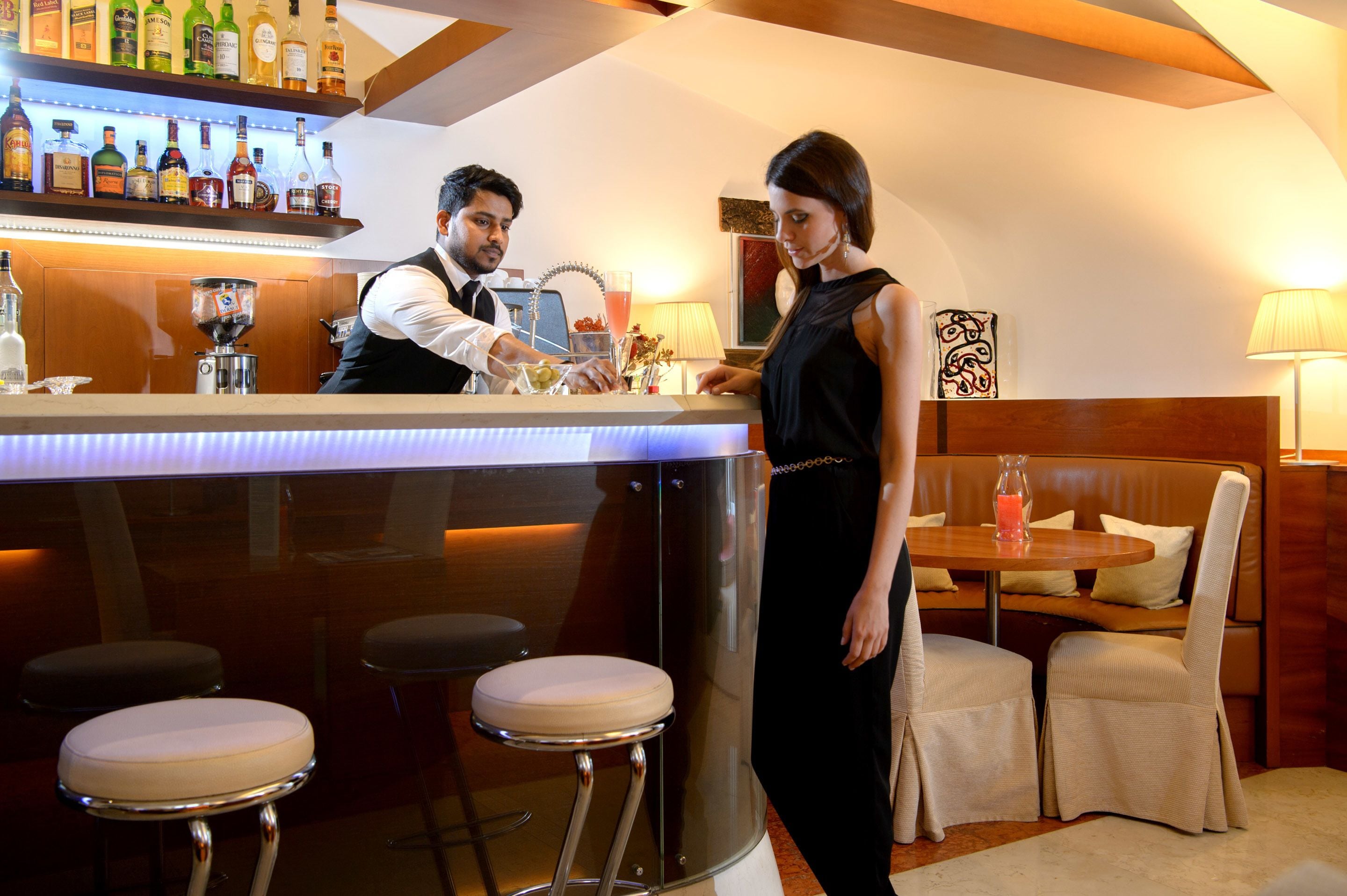 Uma área de bar com banquetas, um barman e uma mulher em pé. Há garrafas nas prateleiras atrás do balcão.
