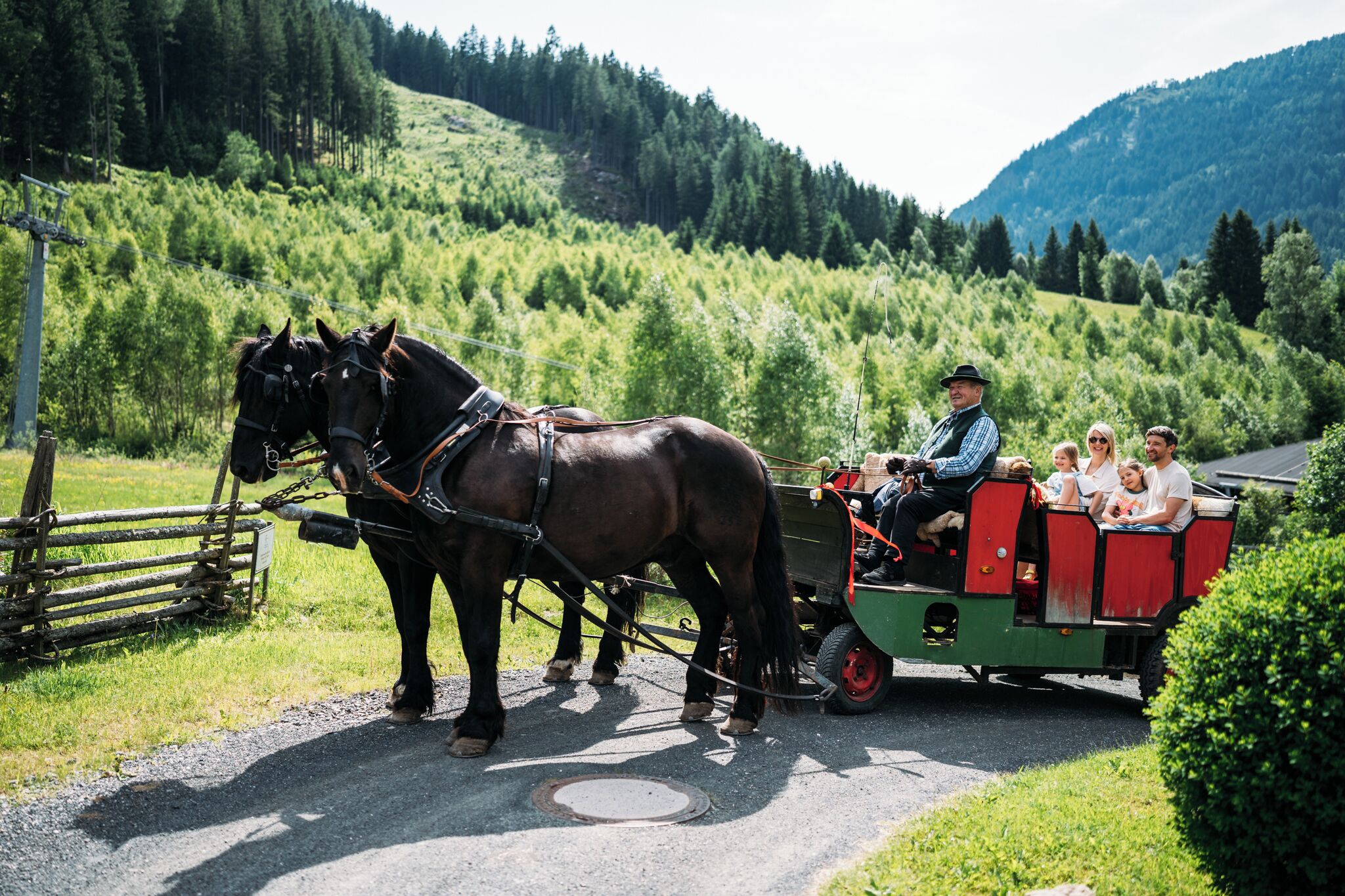 Uma carruagem puxada a cavalo transportando passageiros numa estrada rodeada por vegetação e colinas.