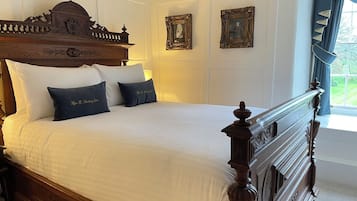 Uma cama clássica de dossel de madeira com roupa de cama branca e almofadas azul-marinho, em frente a uma parede cor de creme com duas molduras de quadro ornamentadas.