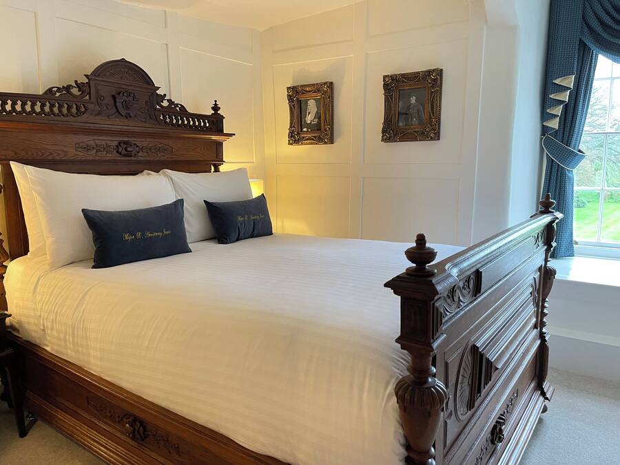 Uma cama clássica de dossel de madeira com roupa de cama branca e almofadas azul-marinho, em frente a uma parede cor de creme com duas molduras de quadro ornamentadas.