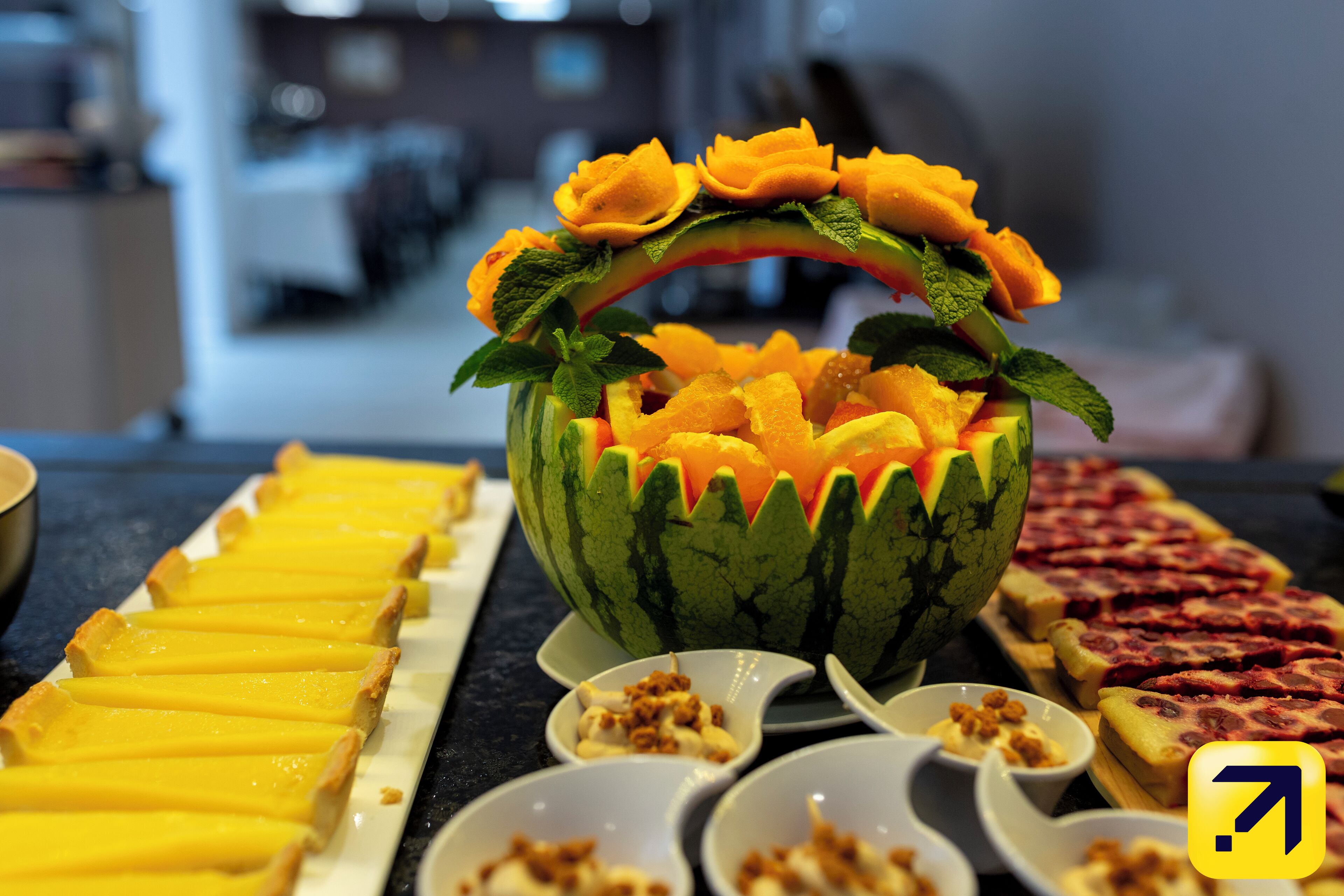 Uma mesa de sobremesas com uma melancia esculpida em flor, uma fileira de tortas de limão e uma seleção de bolos e doces.