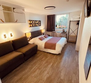 Quarto de hotel com cama, sofá, escrivaninha e janela com vista para árvores.