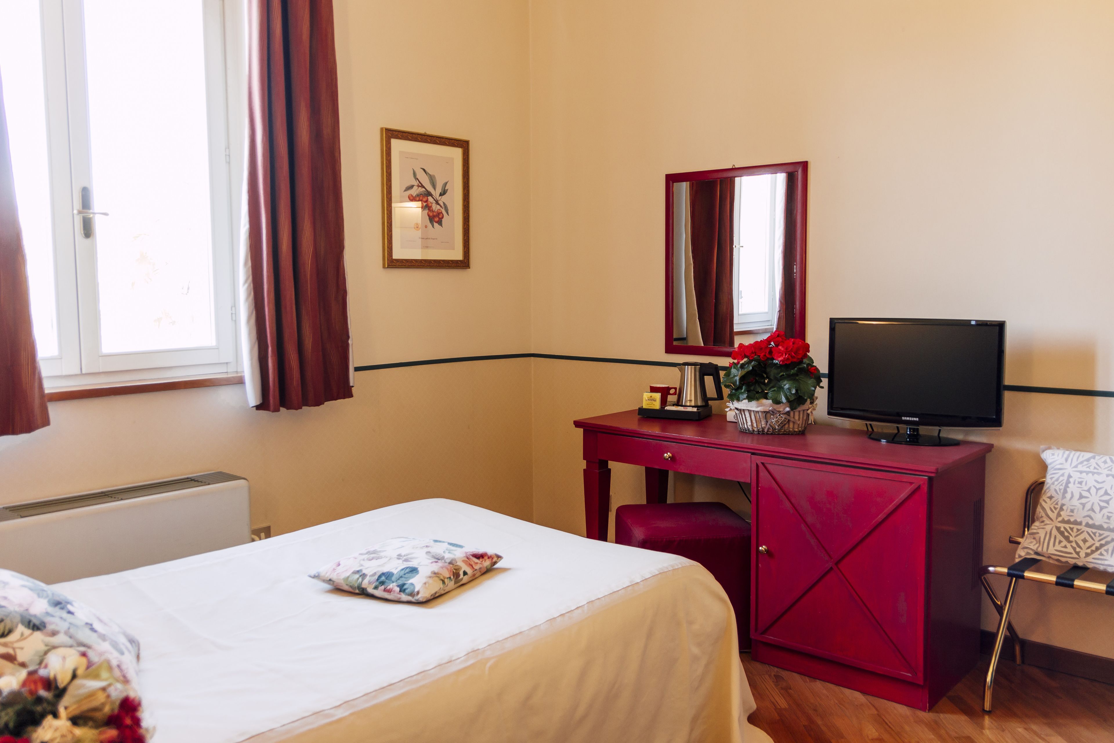 Quarto de hotel com cama, escrivaninha com televisão, armário vermelho e janela com cortinas.