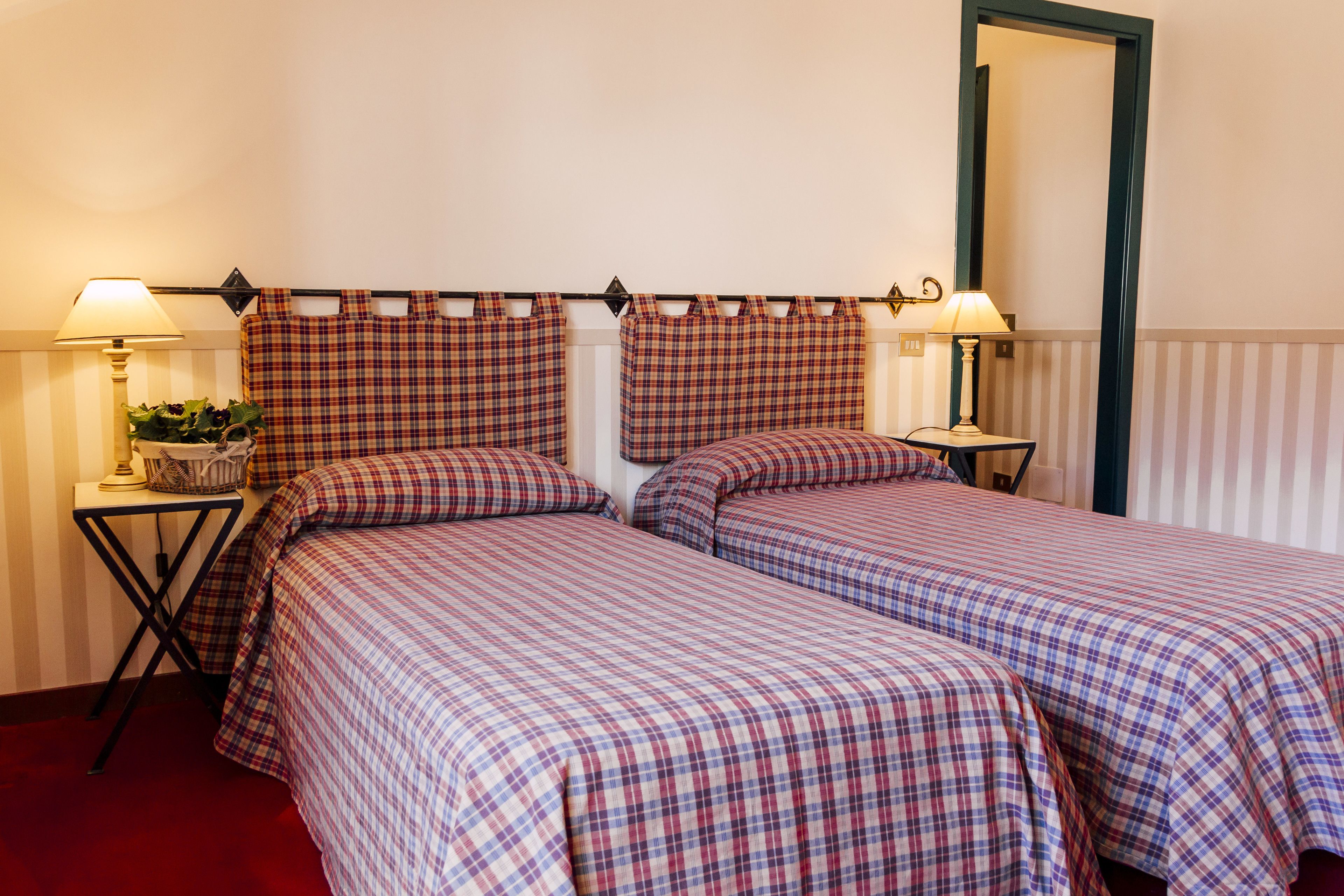 Quarto de hotel com duas camas, cada uma com colcha xadrez, mesa de cabeceira com abajur e planta em vaso.