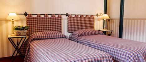 Quarto de hotel com duas camas, cada uma com colcha xadrez, mesa de cabeceira com abajur e planta em vaso.