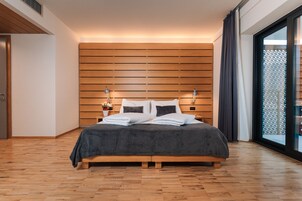 Quarto moderno com cabeceira de madeira, cama grande com almofadas, luminárias laterais e vista através de uma porta de vidro.