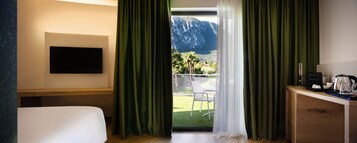 Quarto de hotel com uma cama, uma mesa com cafeteira e vista para uma montanha e jardim através de cortinas translúcidas.