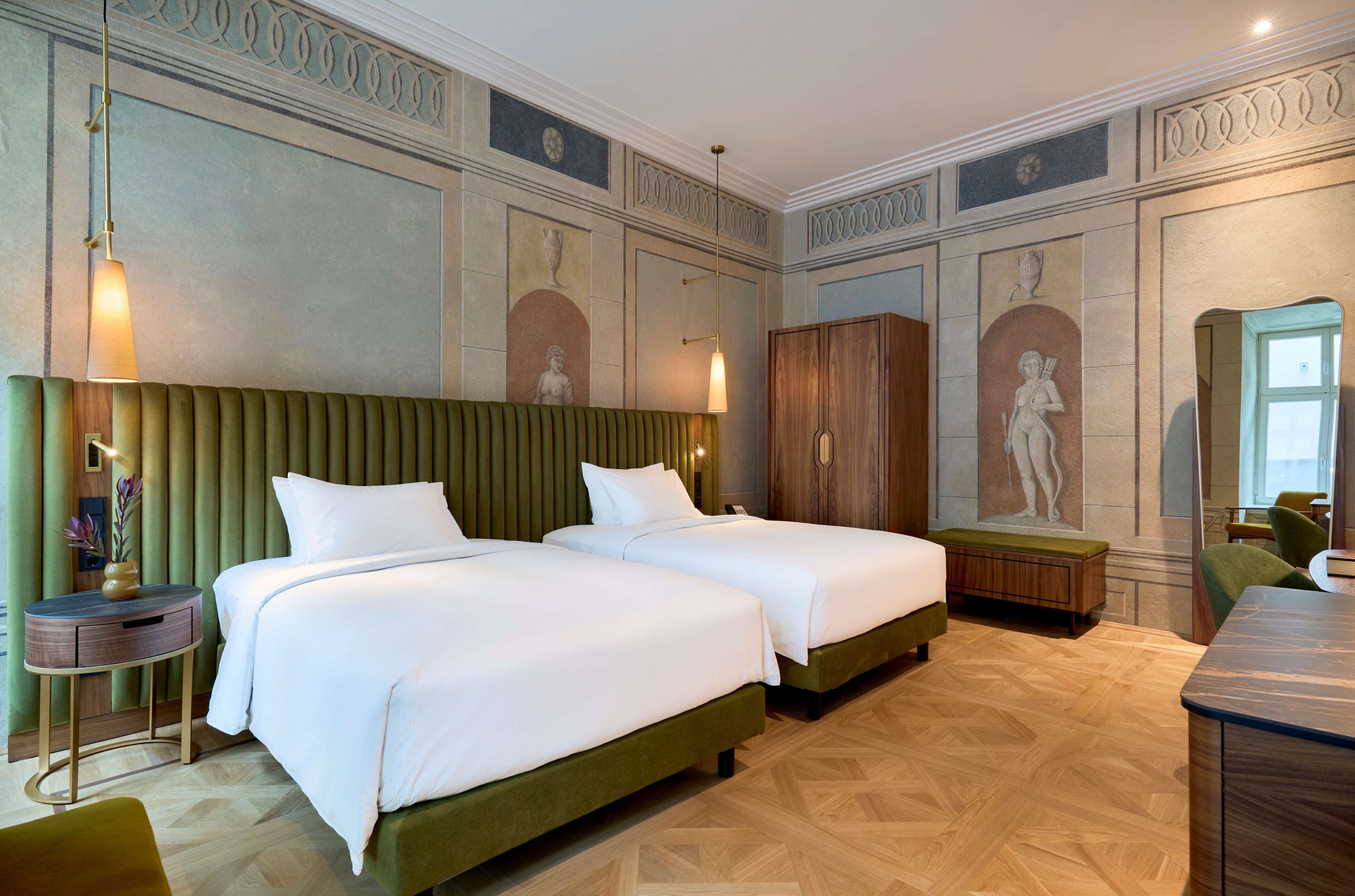 Quarto de hotel com duas camas, um guarda-roupa de madeira, uma mesa lateral redonda e um espelho.