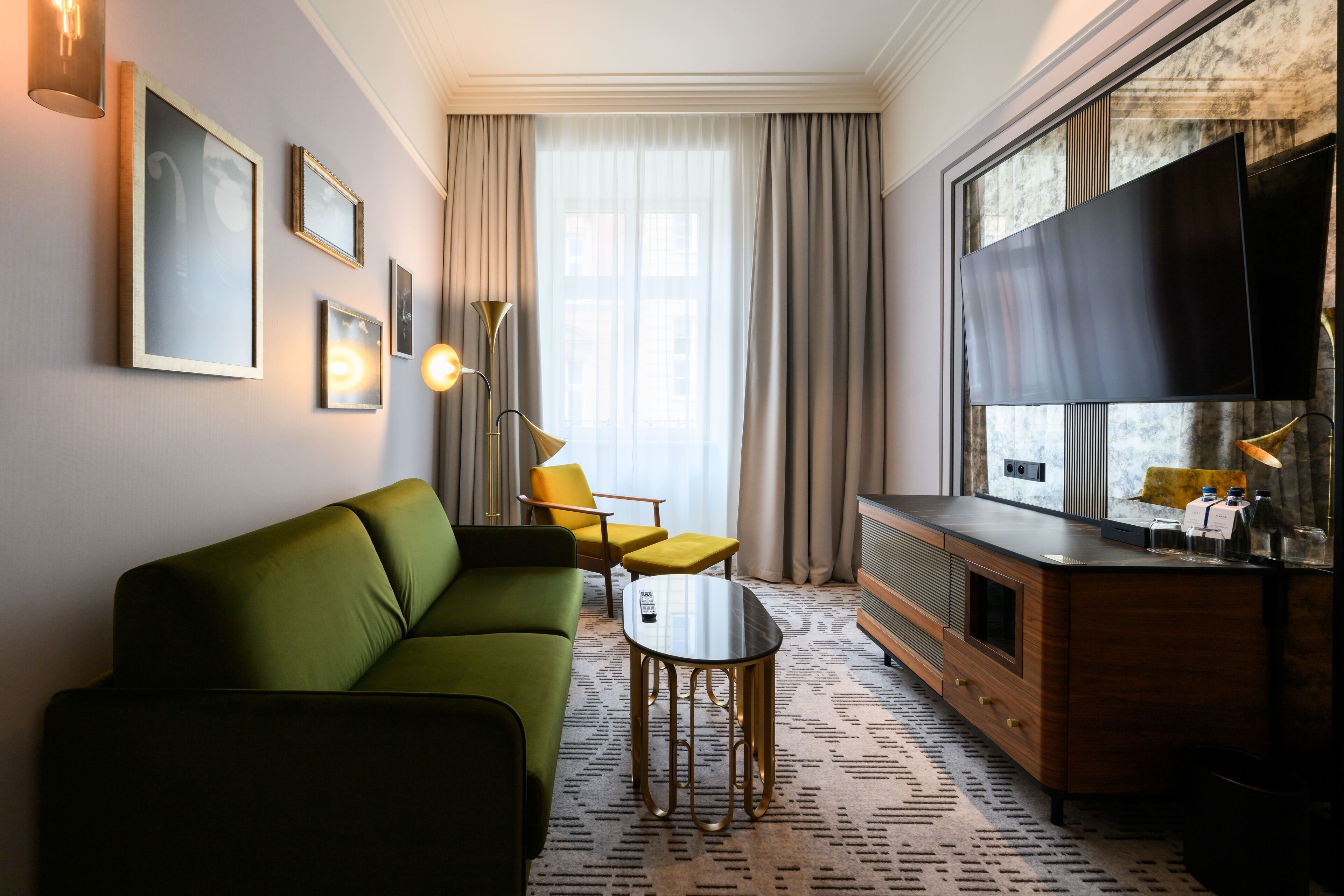 Quarto de hotel moderno com sofá verde, uma mesinha redonda, poltrona amarela, televisão de tela grande e ampla janela com cortinas.