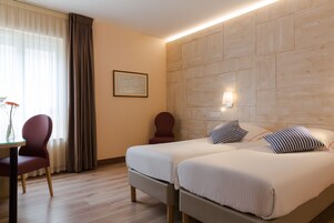 Quarto de hotel com duas camas, uma escrivaninha, uma cadeira e uma janela com cortinas.