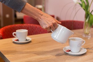 Uma mão servindo café numa xícara sobre uma mesa de madeira, com outra xícara e pires ao fundo.