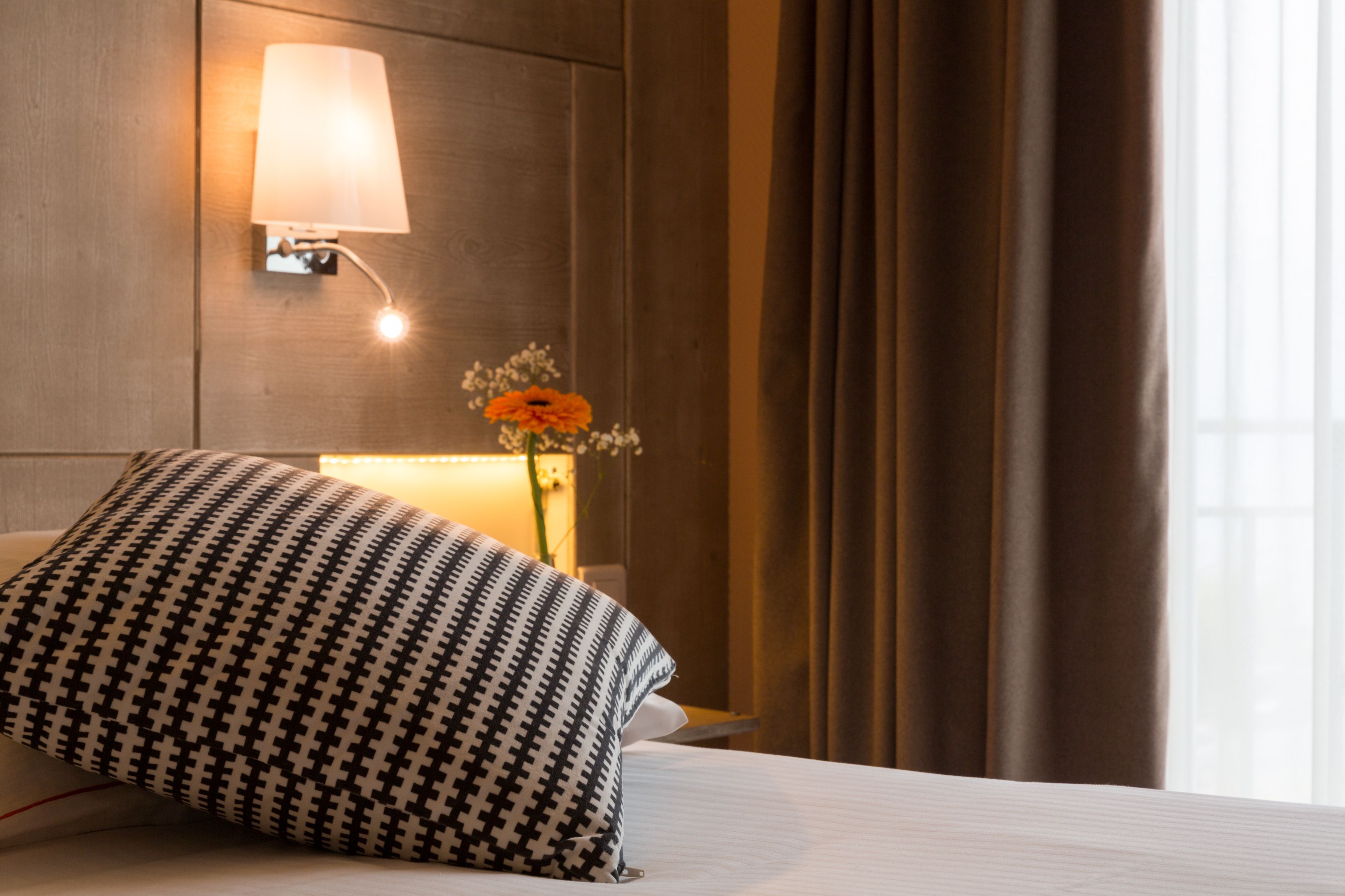 Quarto de hotel com cama, almofada estampada, luminária na parede, um vaso com flores e janela com cortinas.