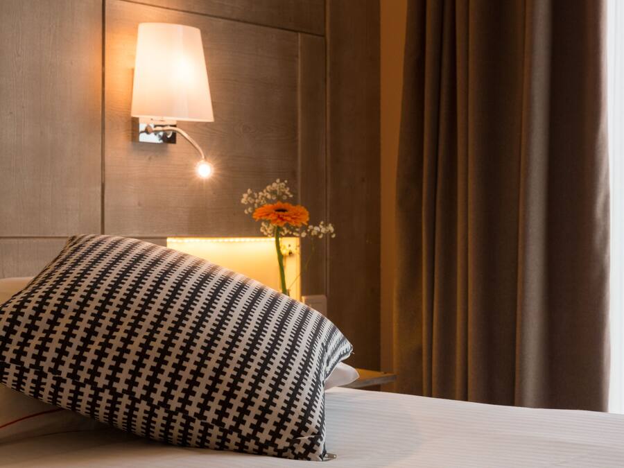 Quarto de hotel com cama, almofada estampada, luminária na parede, um vaso com flores e janela com cortinas.