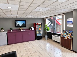 Uma área de recepção com TV de tela plana, refrigerador Coca-Cola, uma planta e uma janela com vista para um edifício.