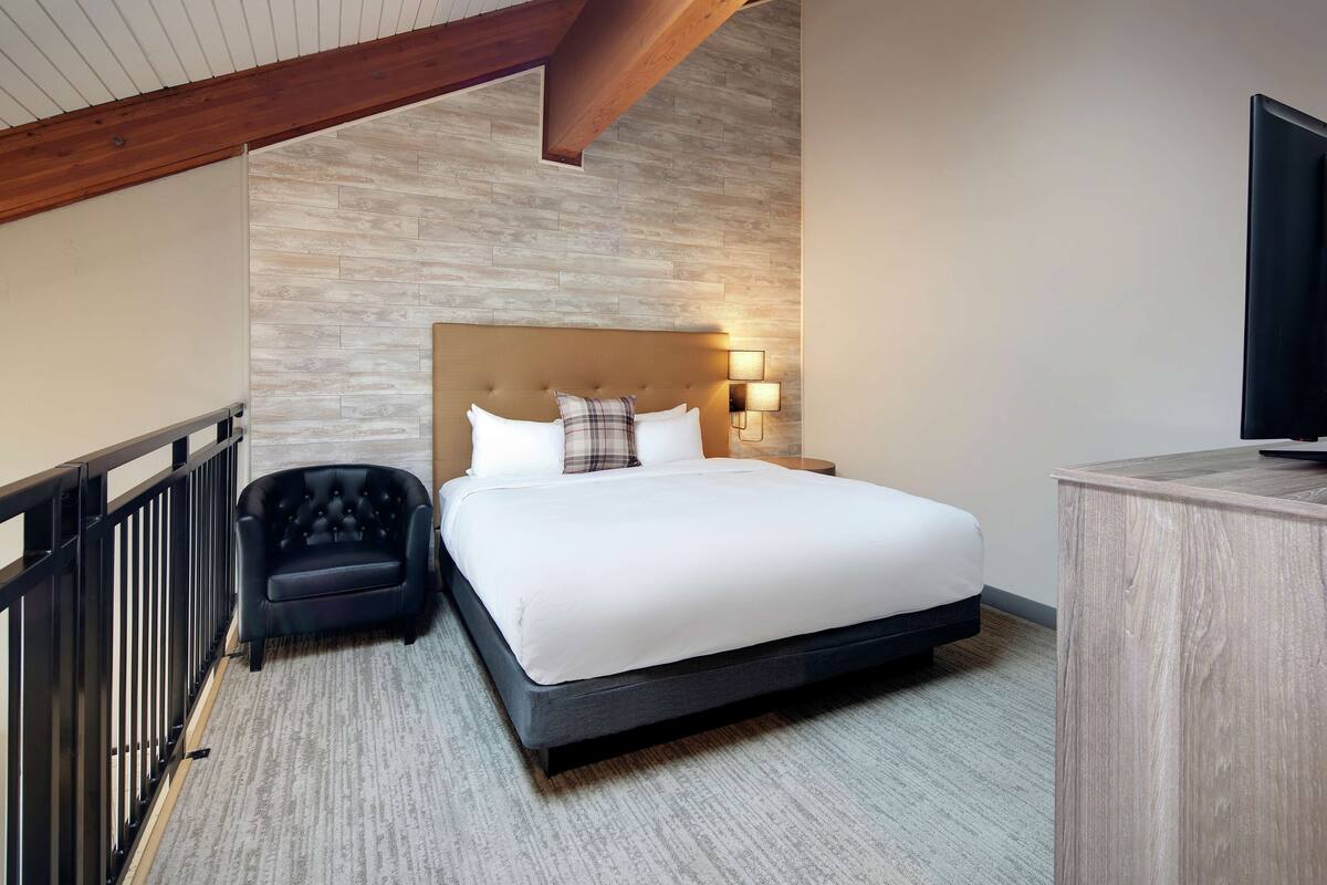 Quarto de hotel com uma cama grande, uma poltrona preta, cabeceira de madeira, televisão e parede de pedra.