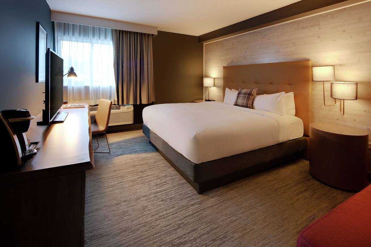 Quarto de hotel com uma cama grande, uma escrivaninha com computador, uma televisão e uma luminária.