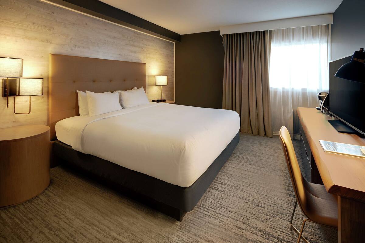 Quarto de hotel com uma cama grande, abajures, uma escrivaninha com televisão e uma janela com cortinas.