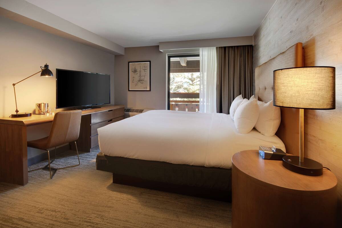 Quarto de hotel com uma cama grande, uma escrivaninha com computador, uma televisão e um abajur.