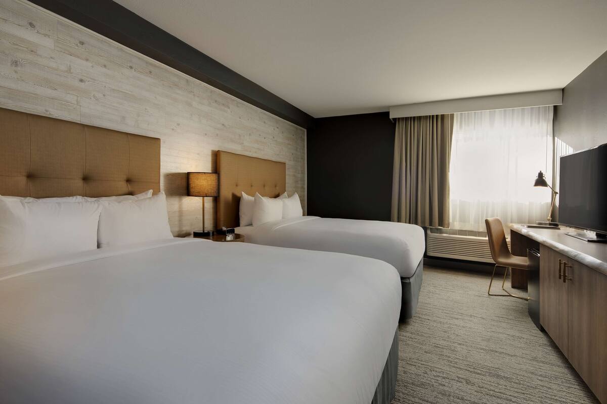 Quarto de hotel com duas camas grandes, uma escrivaninha com cadeira, televisão e janela com cortinas.