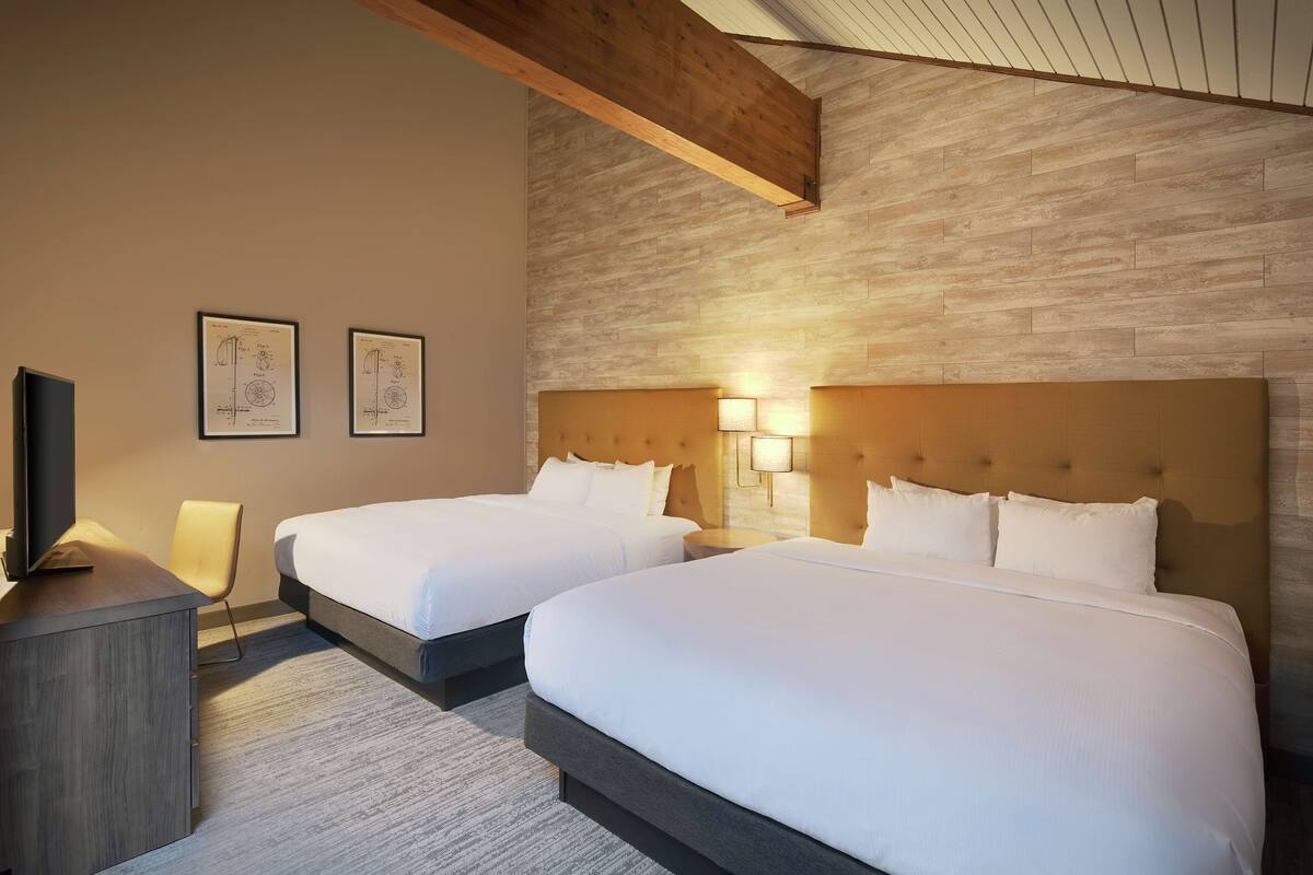 Quarto de hotel com duas camas, televisão, escrivaninha e diplomas emoldurados na parede.